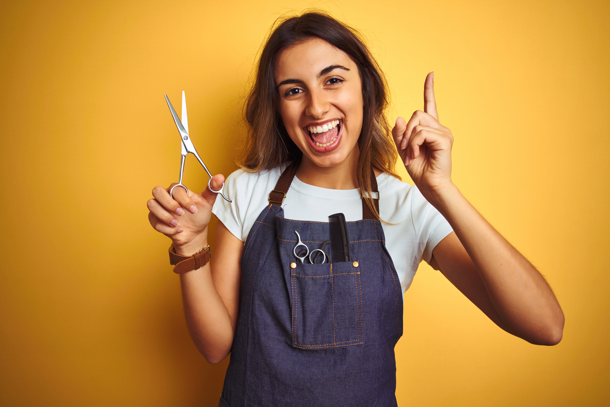 Young beautiful hairdresser woman holding scissors over yellow isolated ...
